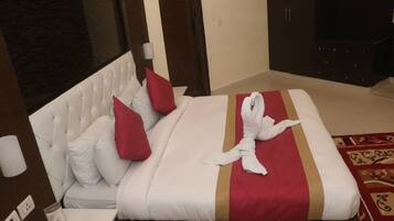 Quarto de hotel com uma cama grande, roupa de cama branca, almofadas vermelhas, edredom vermelho e dourado, toalha branca sobre a cama, mesa de cabeceira com controle remoto e luminária fixada na parede.