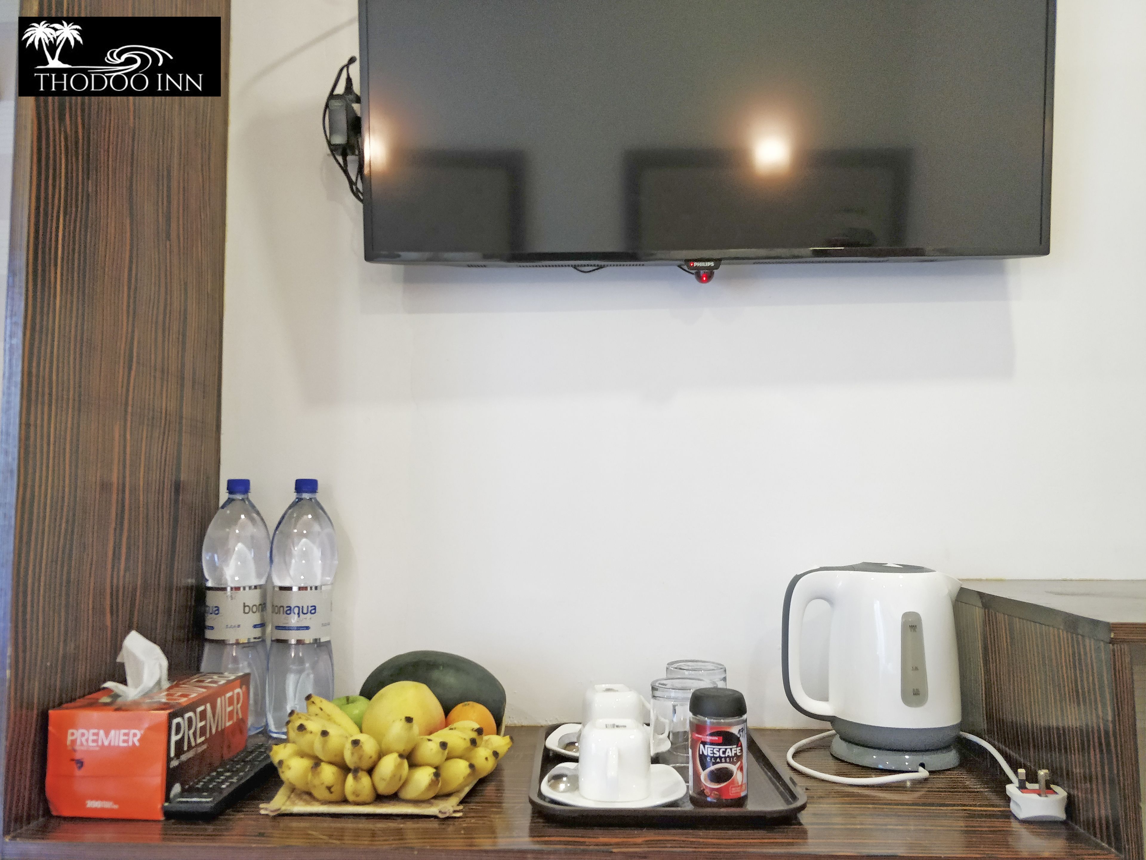 Quarto de hotel com televisão de ecrã plano na parede, minibar com água engarrafada, cesta de frutas, máquina de café e chaleira.