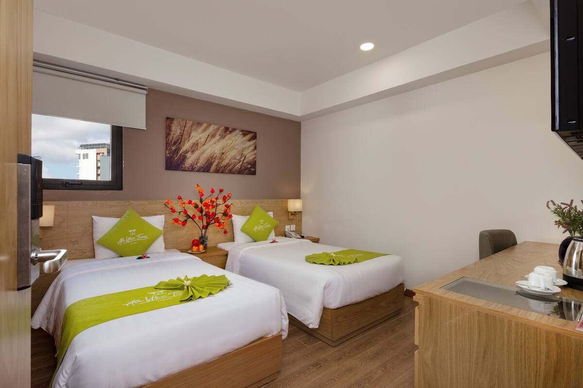 Quarto de hotel com duas camas, uma escrivaninha, um espelho e uma janela com vista para edifícios.