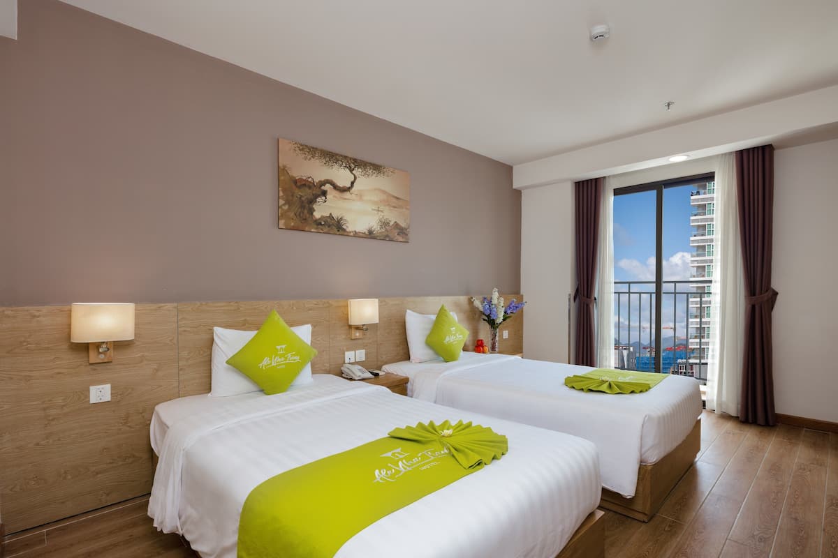 Quarto de hotel com duas camas, um quadro na parede, vista da cidade pela janela e uma varanda com cortinas.