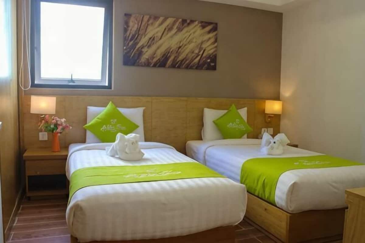 Quarto de hotel com duas camas, uma janela, um quadro na parede e uma mesa de cabeceira com um vaso de flores.
