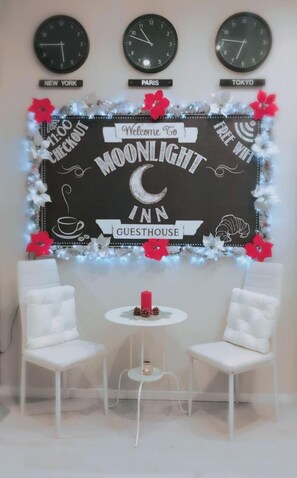 Paredes com relógios indicando 'Nova York', 'Paris' e 'Tóquio', uma placa com a inscrição 'Bem-vindo(a) à Pousada Moonlight Inn', e duas cadeiras brancas com uma pequena mesa entre elas.