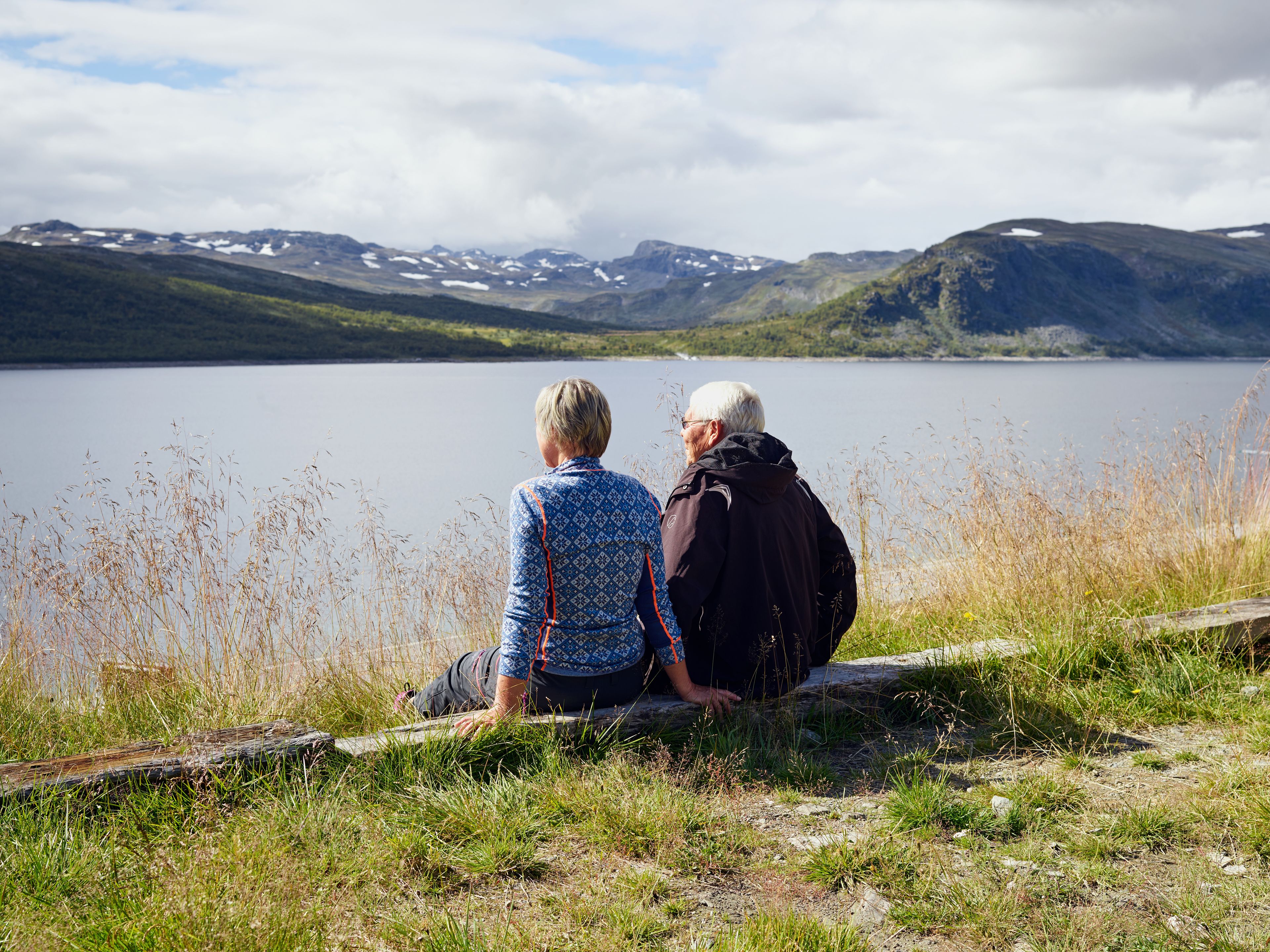 Duas pessoas sentadas em uma rocha à beira de um lago, com montanhas ao fundo.