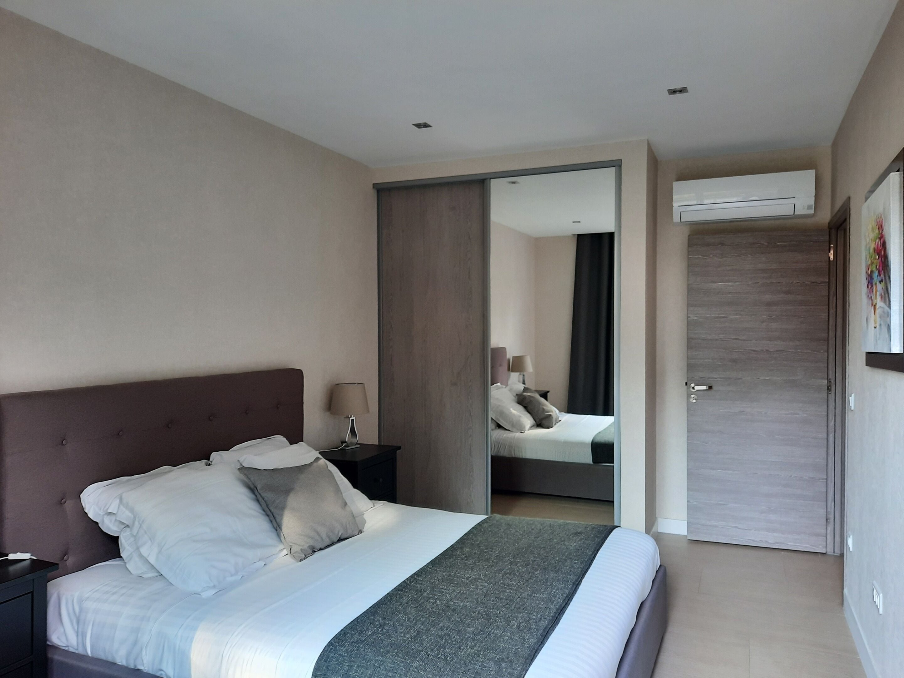 Quarto de hotel moderno com uma cama grande, mesa de cabeceira, espelho e aparelho de ar condicionado.