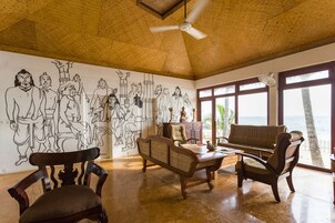 Quarto com teto alto, ventoinha no teto, um grande mural na parede, um sofá, duas poltronas e uma mesa de madeira.