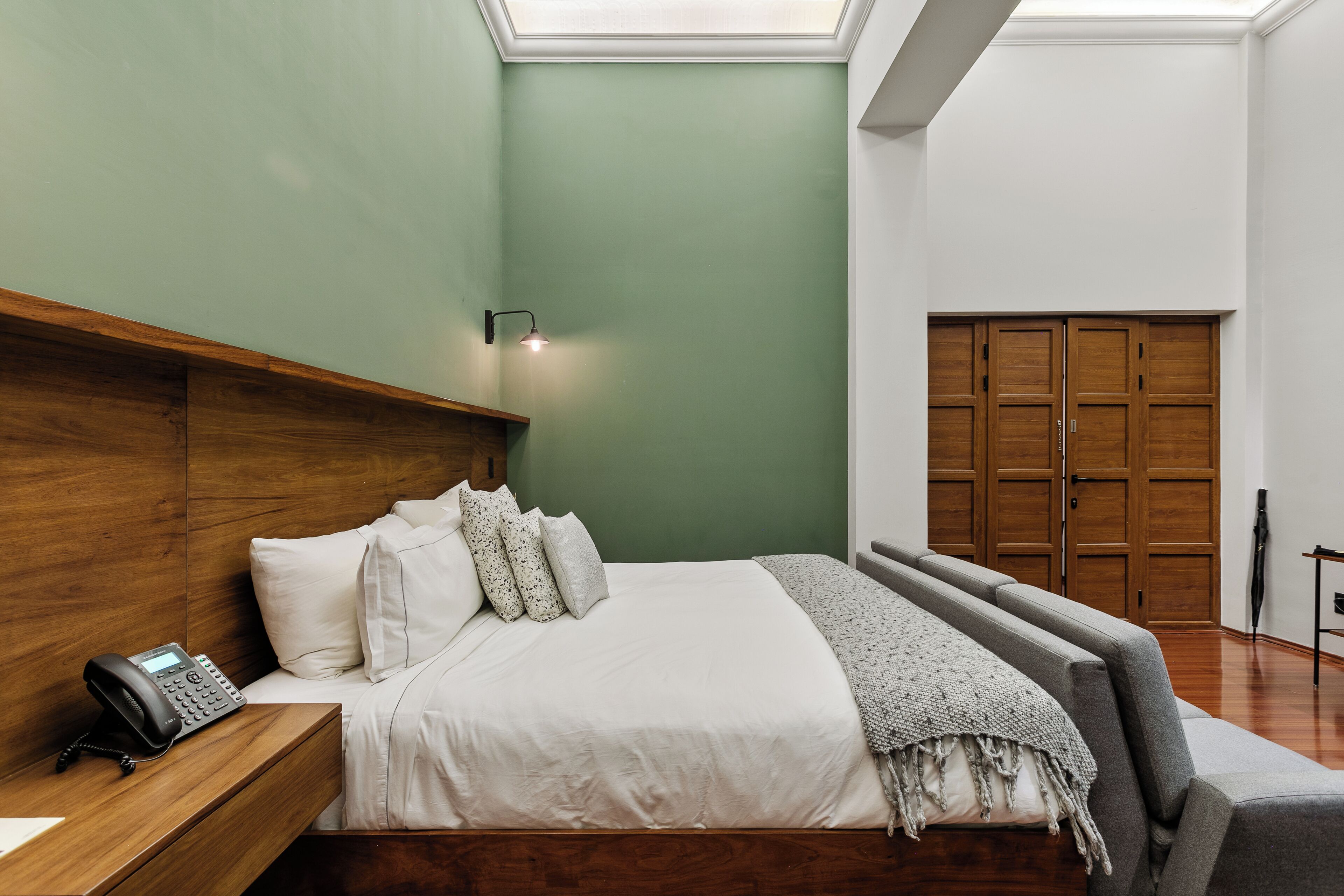 Quarto de hotel com cabeceira de madeira, cama com roupa de cama branca, telefone ao lado da cama e vista para uma porta de madeira e corredor.
