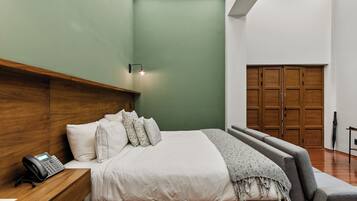 Quarto de hotel com cabeceira de madeira, cama com roupa de cama branca, telefone ao lado da cama e vista para uma porta de madeira e corredor.