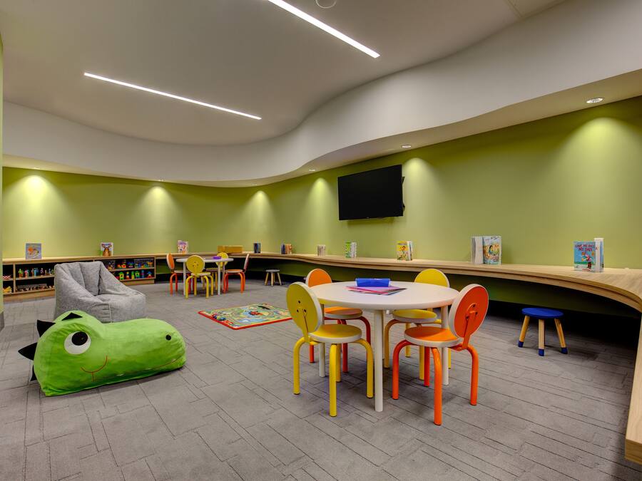 Uma biblioteca infantil com cadeiras coloridas, um pufe em formato de feijão verde e uma televisão de tela plana fixada na parede.