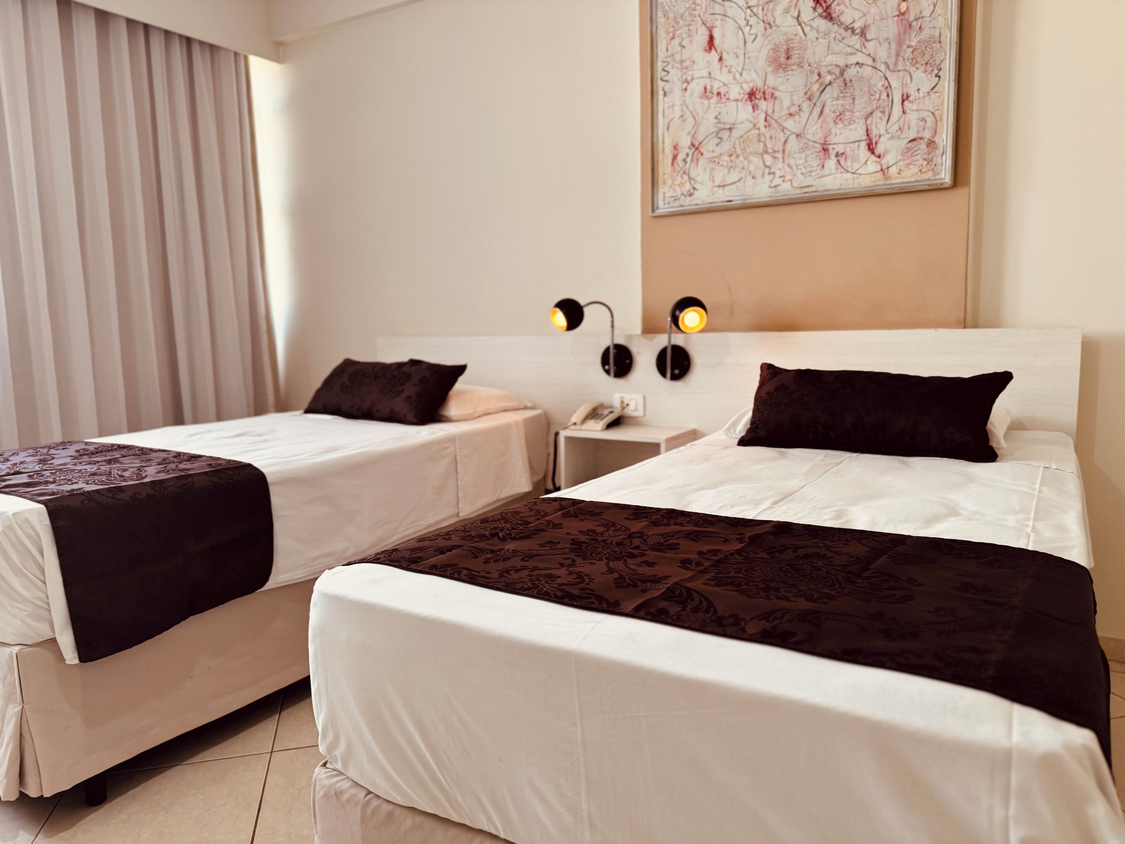 Quarto de hotel com duas camas de solteiro, uma mesinha de cabeceira com telefone, duas luminárias fixadas na parede e um quadro abstrato grande na parede.