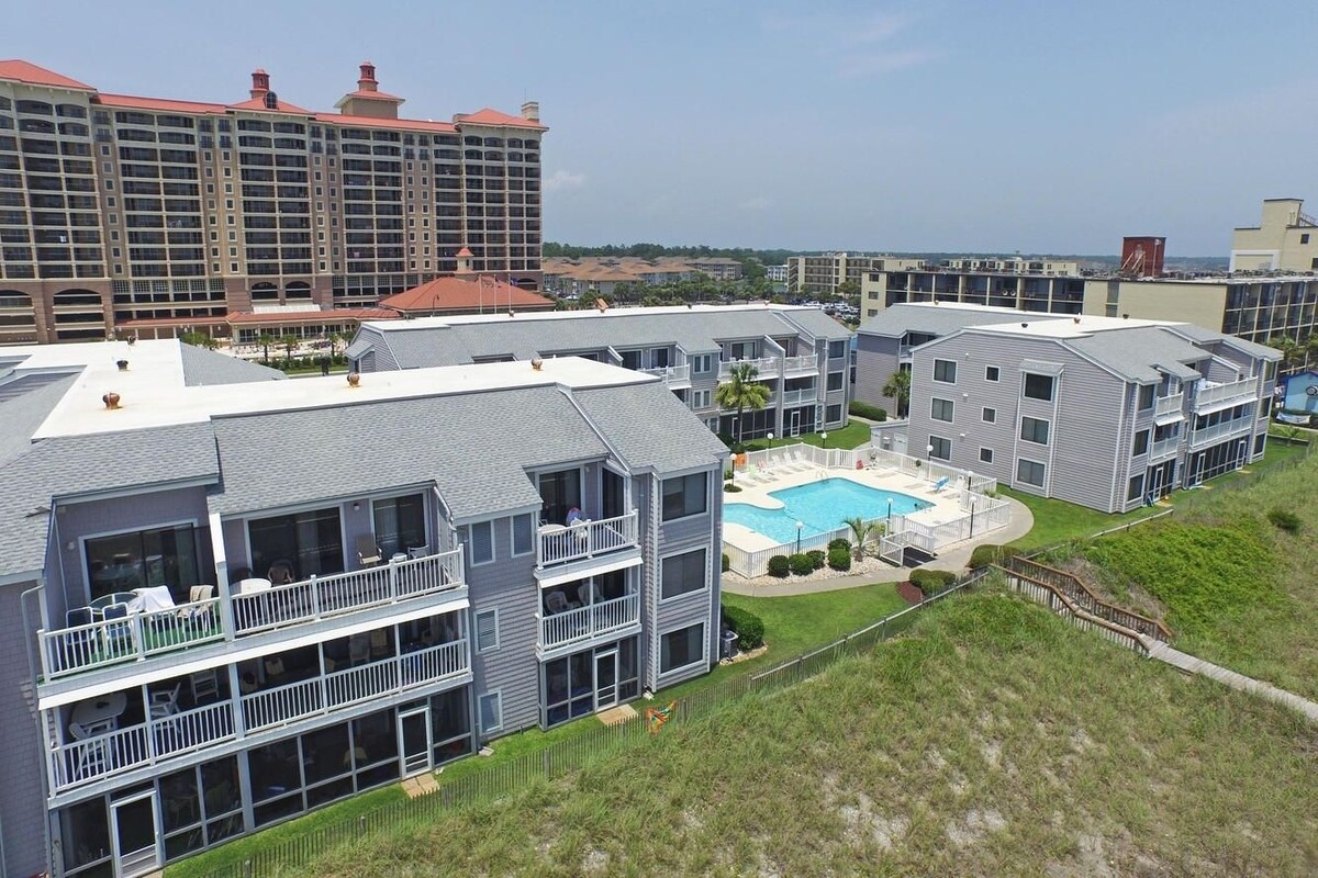 Um edifício residencial com vários andares, piscina no terraço e um prédio grande ao fundo.