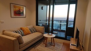 Uma sala de estar com um sofá bege, uma mesa redonda pequena, um rack de TV e uma janela grande com vista para a cidade.