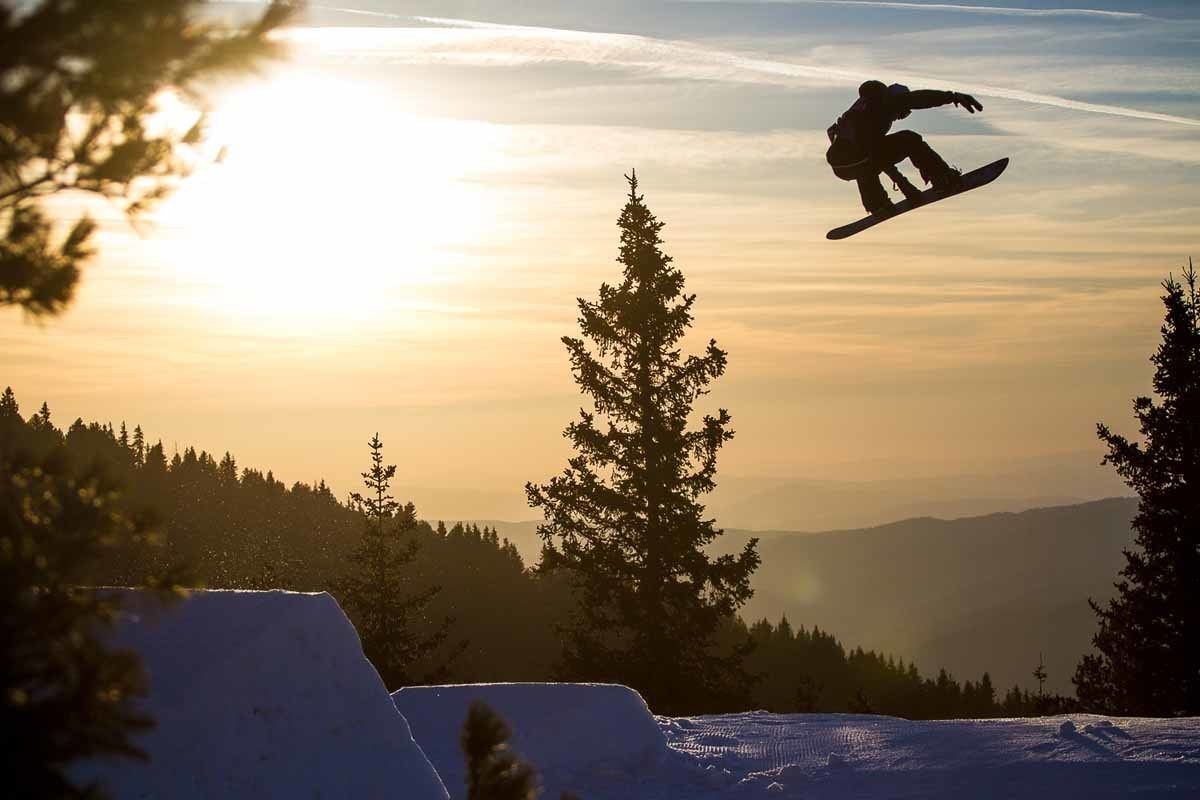 Um snowboarder no ar, com o sol poente ao fundo, terreno coberto de neve e árvores.