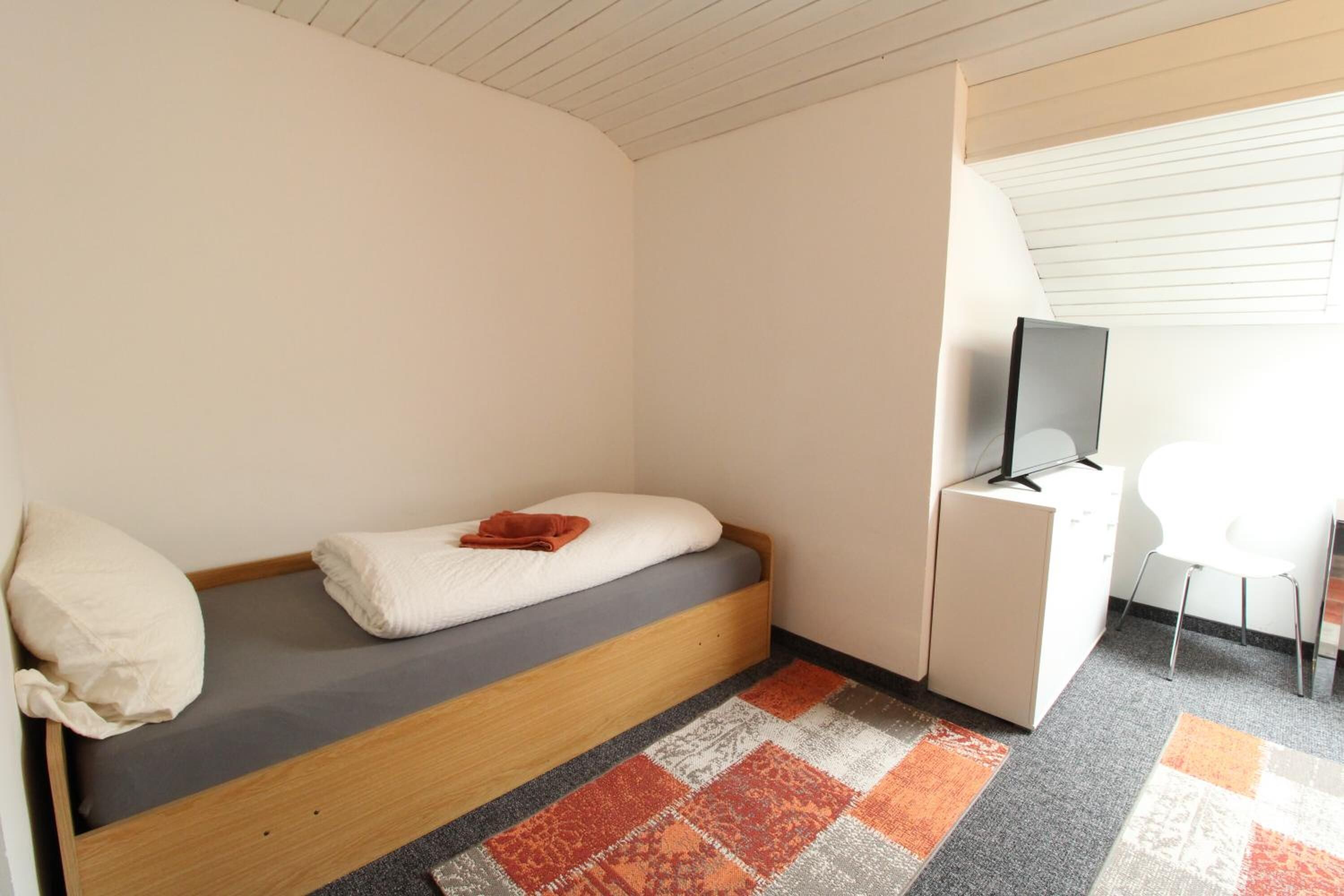 Quarto pequeno com uma cama de solteiro, escrivaninha e cadeira, televisão e tapete estampado.
