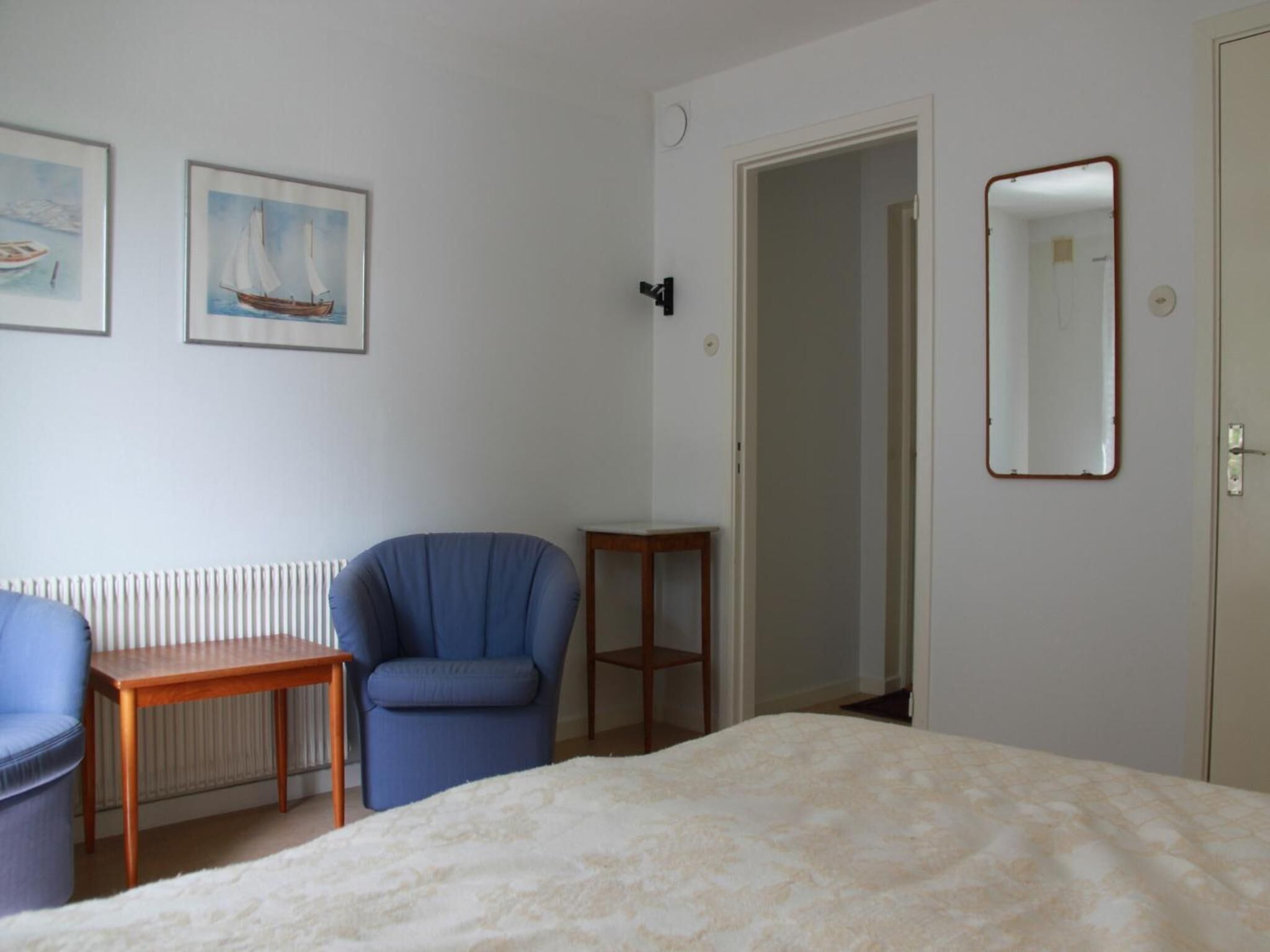 Quarto com cama, uma poltrona azul, uma pequena mesa de madeira e duas pinturas emolduradas na parede.