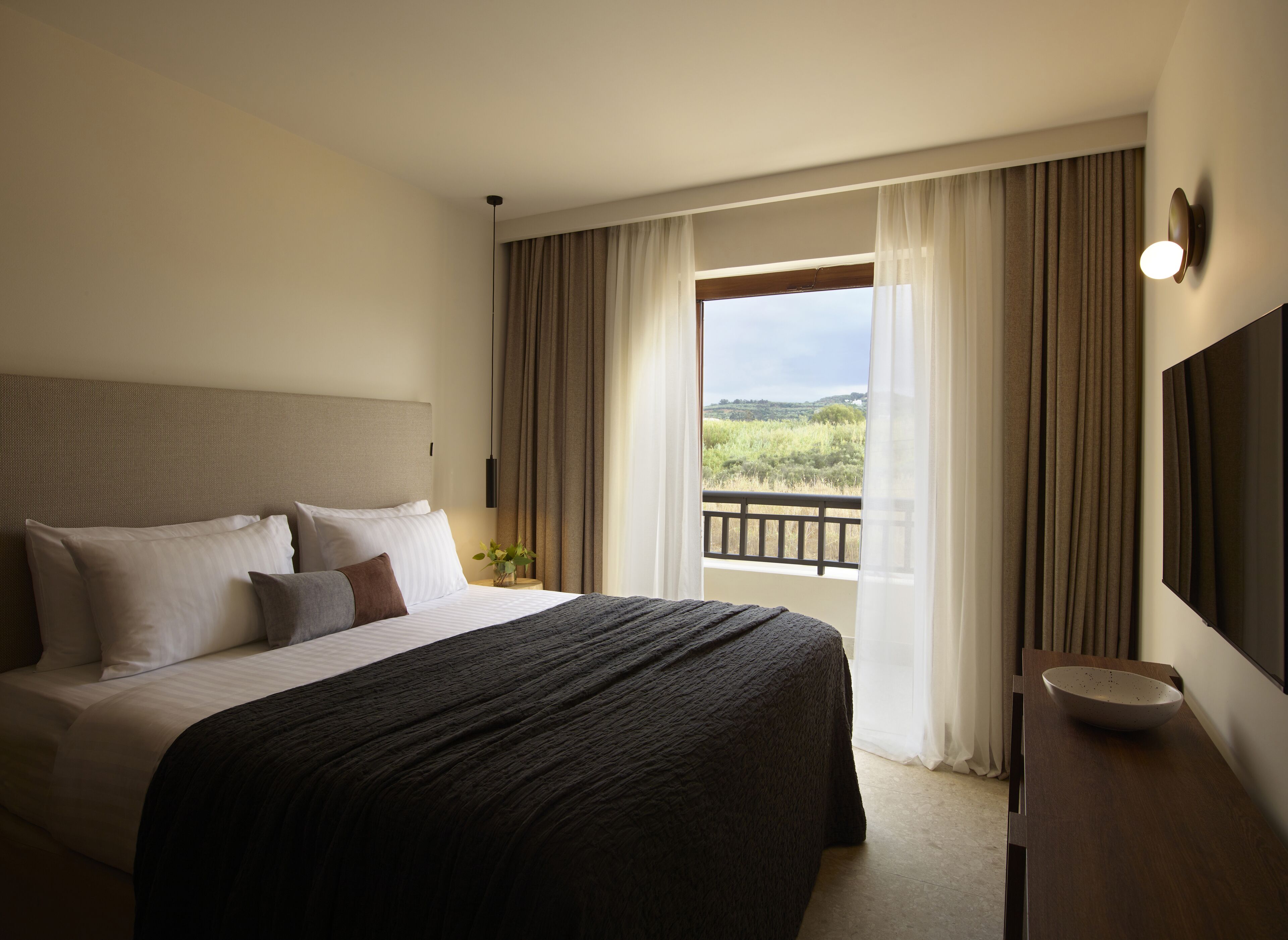 Quarto de hotel com uma cama grande, uma TV sobre uma mesinha de madeira e uma varanda com cortinas.