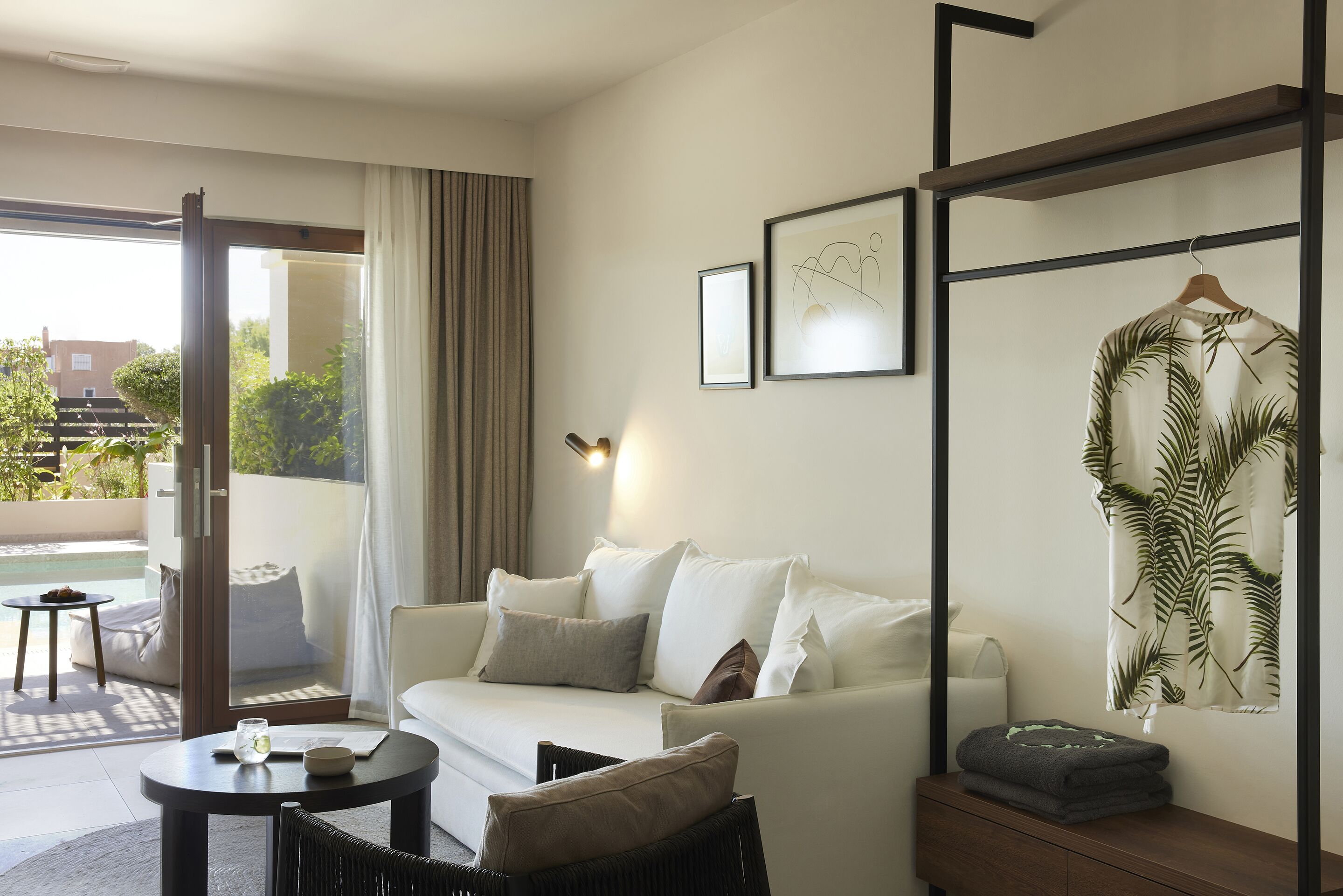 Quarto de hotel moderno com sofá branco, uma mesa redonda pequena, uma cadeira de vime, cômoda de madeira e uma grande janela com vista para vegetação e uma piscina.