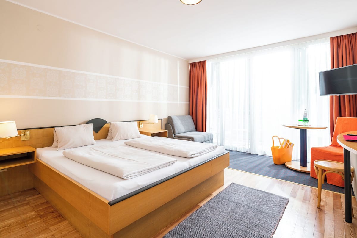Quarto de hotel com uma cama grande, uma escrivaninha, uma cadeira, uma TV e uma janela com cortinas.