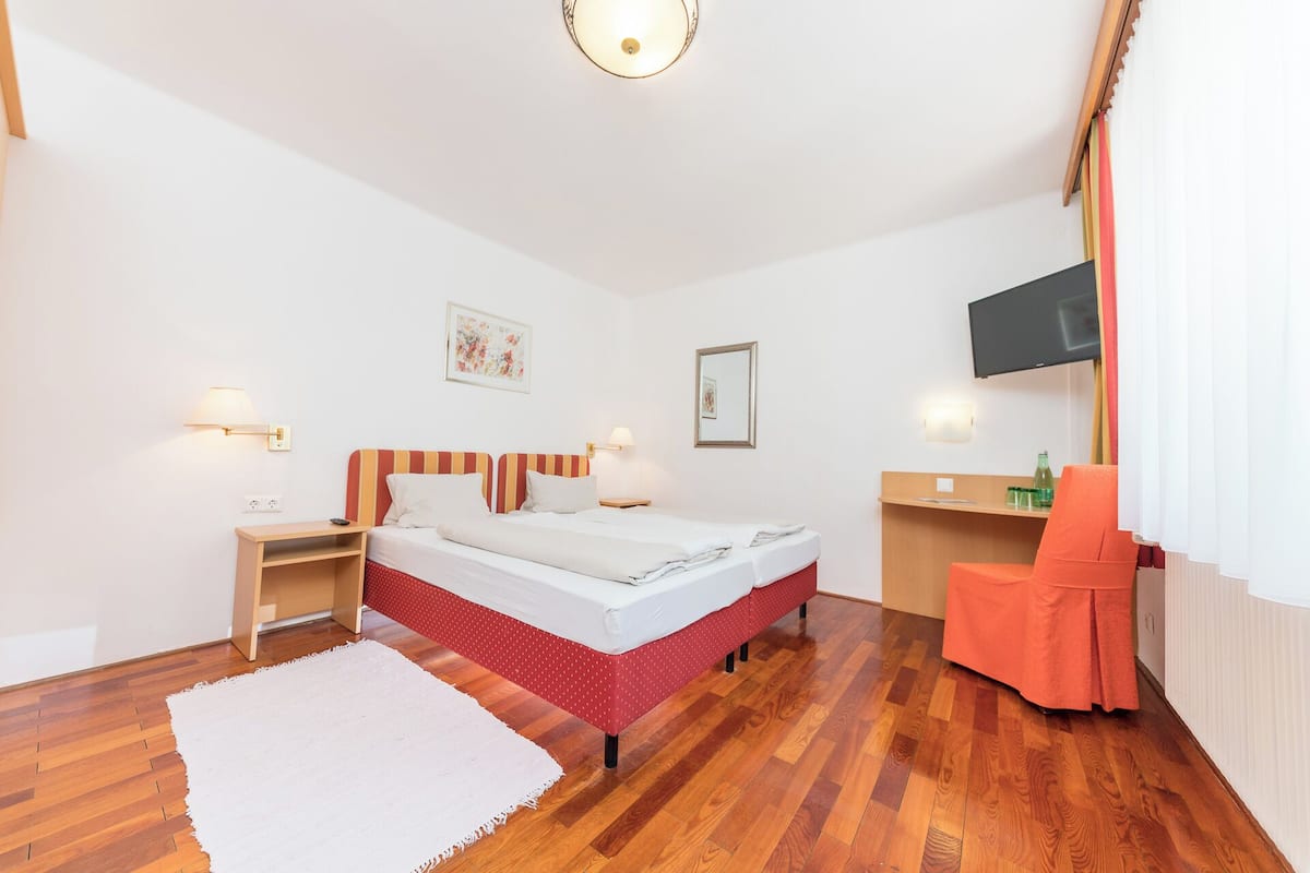Quarto de hotel com cama, mesas de cabeceira, escrivaninha, cadeira, televisão e janela com cortinas.