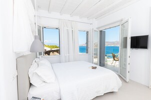 Quarto com uma cama grande, cortinas brancas e vista para o mar através de portas abertas.