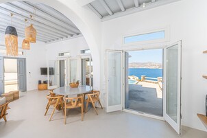 Um quarto espaçoso com uma grande mesa redonda e cadeiras, teto com vigas aparentes e vista para o mar através de uma porta de vidro.