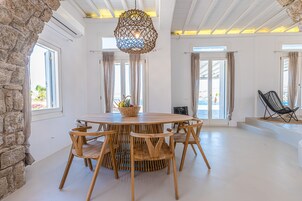 Uma sala de jantar ampla com uma mesa redonda de madeira, rodeada por cadeiras, um lustre pendurado e grandes janelas com cortinas.