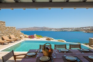 Uma área de refeições junto à piscina, com uma mesa posta para dois, com vista para o mar azul e uma paisagem montanhosa.