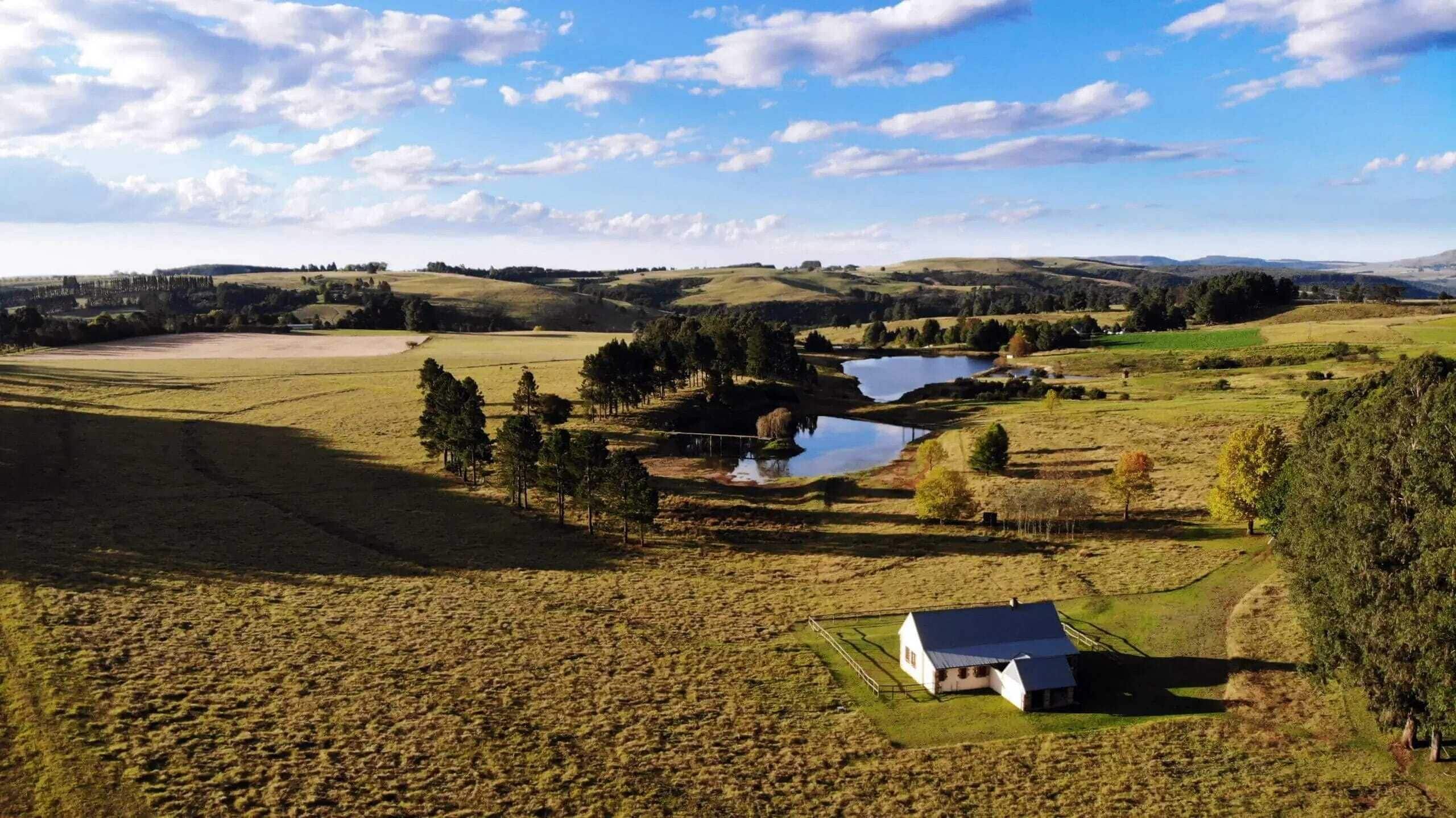 Paisagem rural com um pequeno edifício branco, um lago e colinas suaves ao fundo.