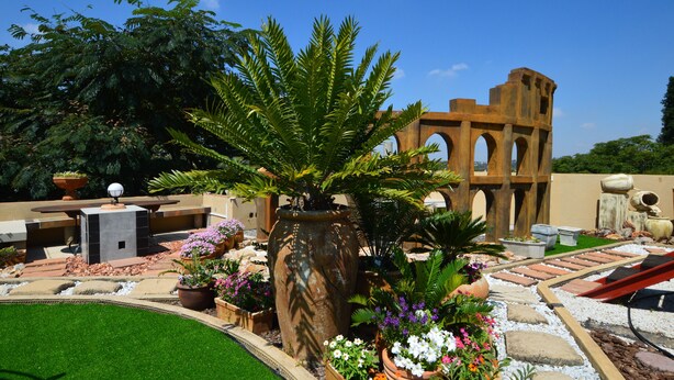 Um jardim ajardinado com uma grande palmeira, diversos vasos de plantas e um edifício com arcos ao fundo.
