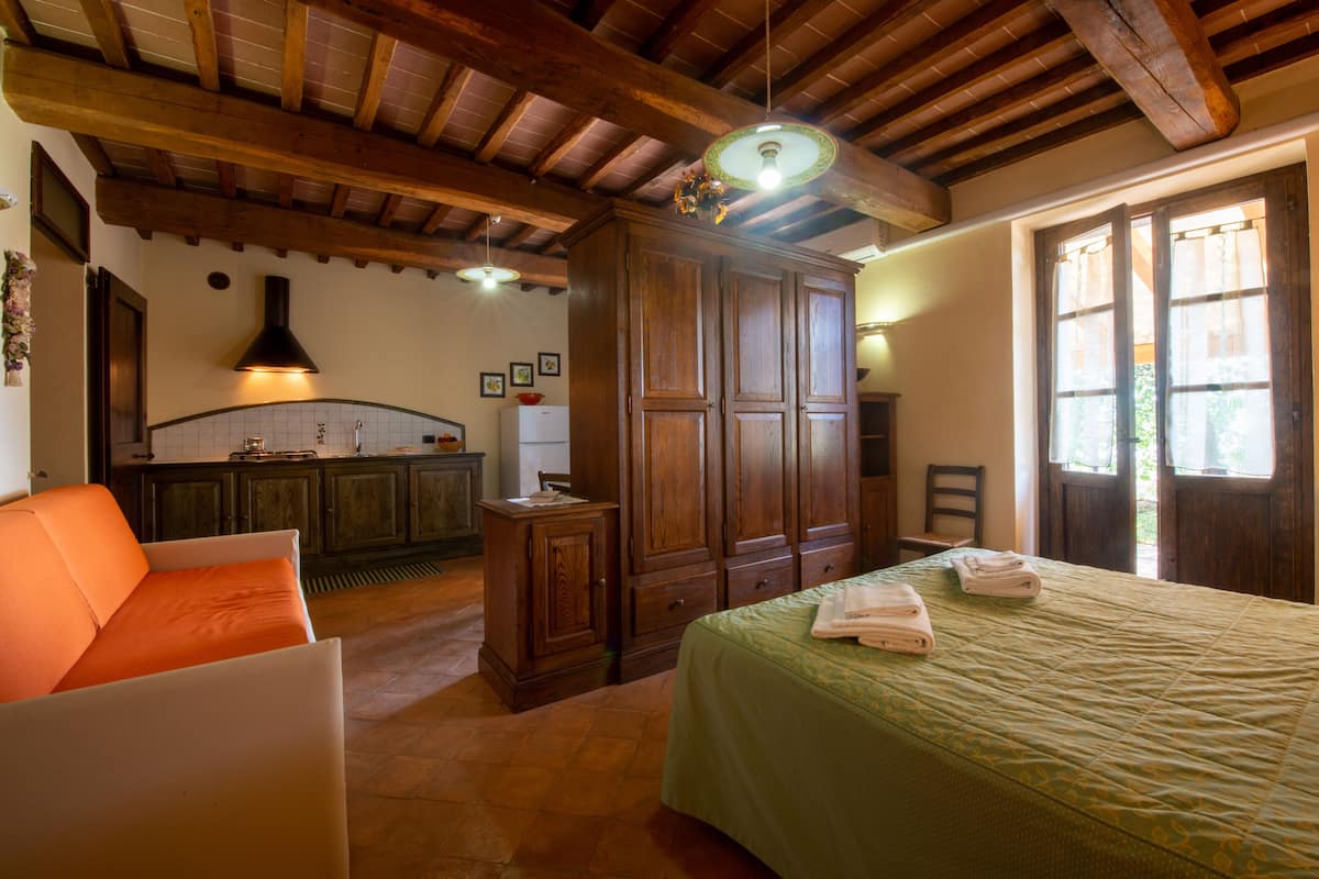 Quarto com cama, sofá, guarda-roupa de madeira, área de cozinha e janela com cortinas.