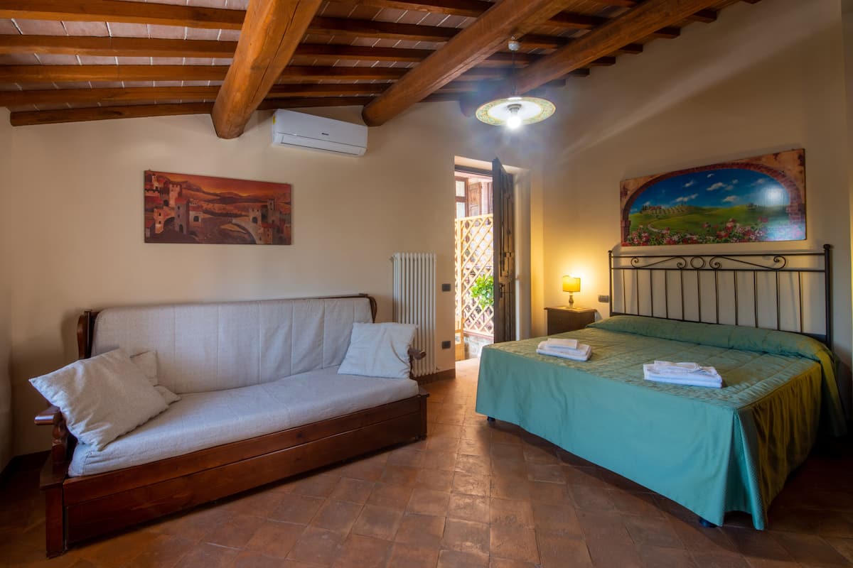 Quarto com teto de madeira, cama com colcha verde, sofá com almofada branca e um quadro na parede.