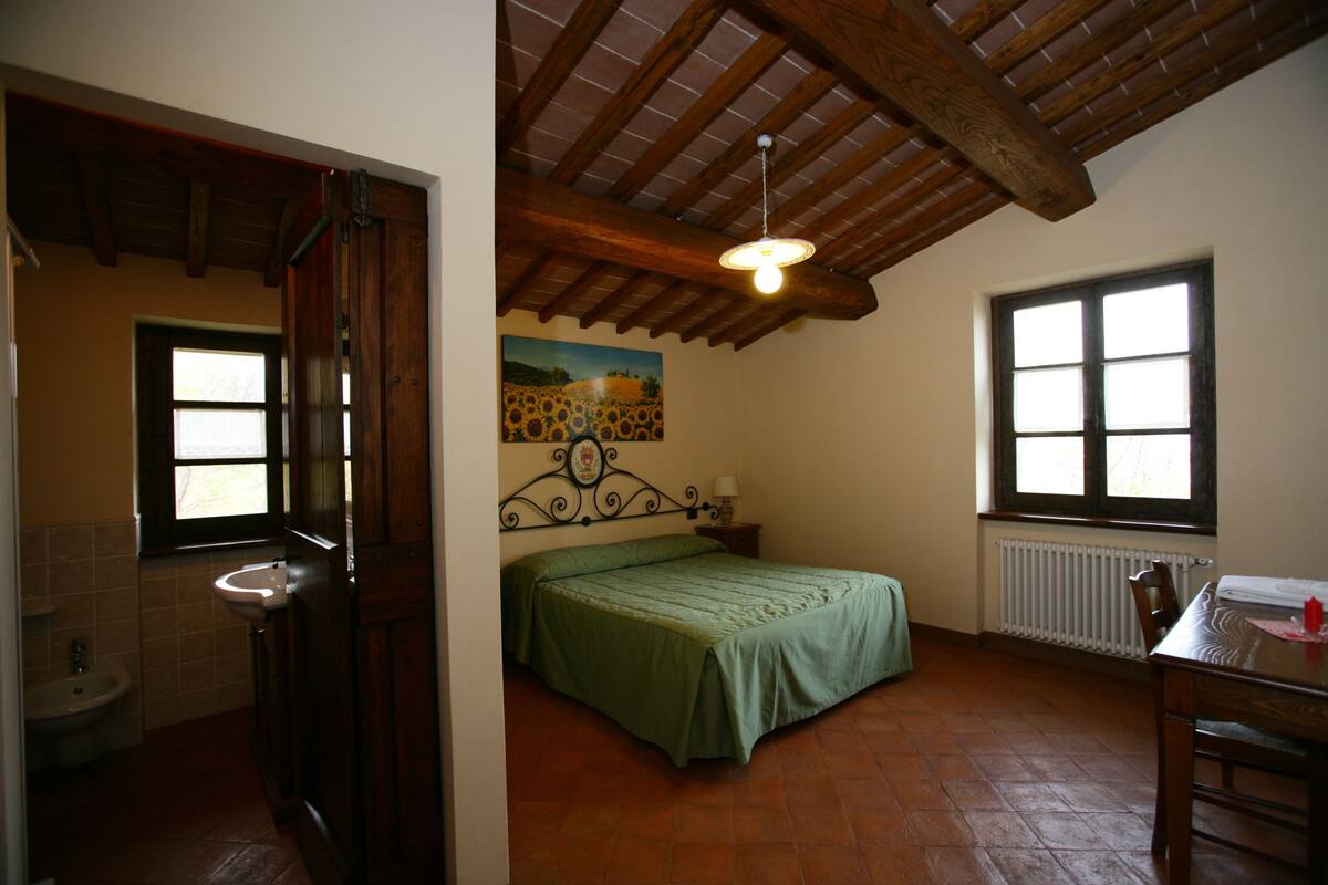 Quarto com teto de madeira, cama com colcha verde, mesa de cabeceira com abajur, janela e radiador.
