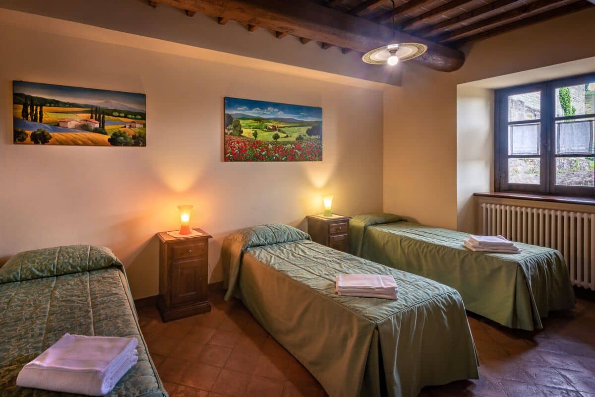 Quarto com duas camas de solteiro, mesa de cabeceira com abajur e janela com vista para vegetação.