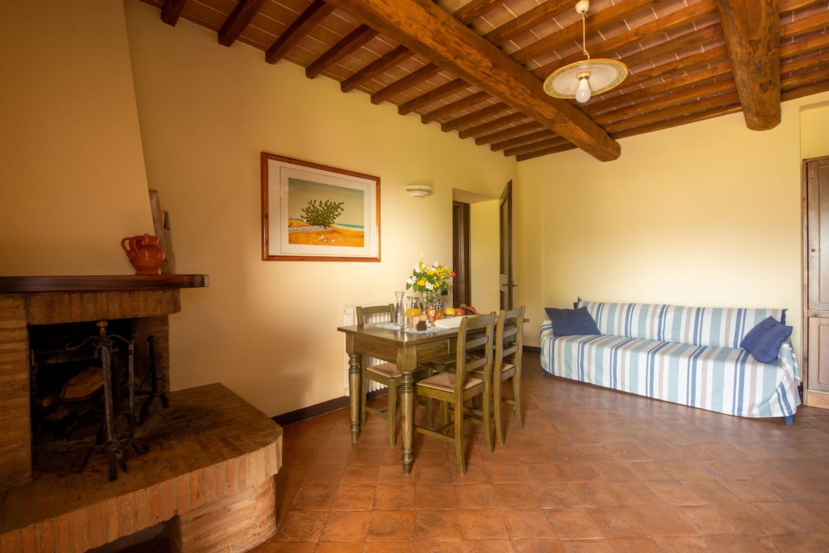 Uma área de refeições com lareira de tijolos, mesa de jantar de madeira e cadeiras. Há um sofá listrado e um quadro na parede.