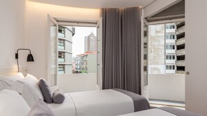 Quarto de hotel moderno com duas camas, uma janela grande com cortinas e vista para a paisagem urbana.