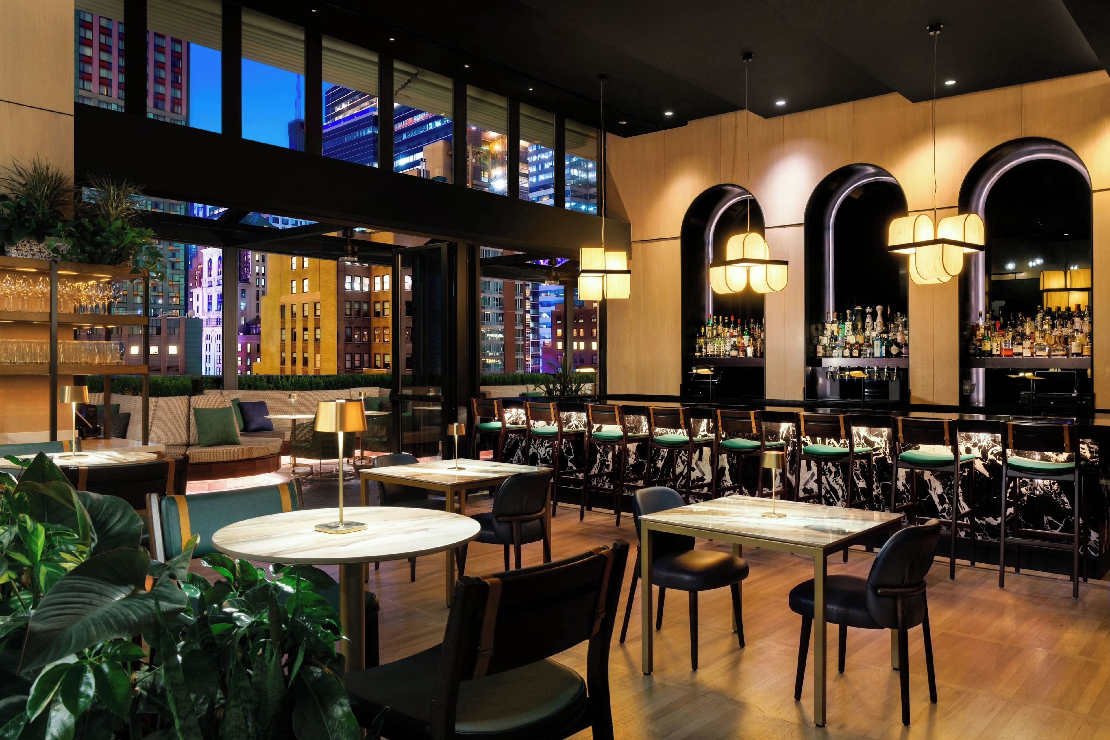 Uma área de bar moderna com mesas redondas, banquetas e vista para o horizonte urbano através de grandes janelas.
