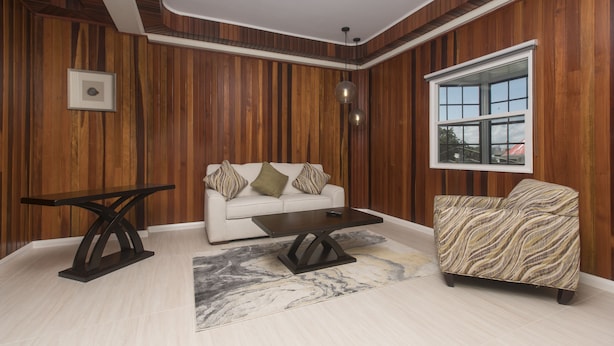Uma sala de estar moderna com painel de madeira, um sofá branco com almofadas estampadas, uma poltrona estampada, uma mesa de centro escura e uma grande janela com vista.