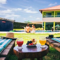 Uma mesa de madeira com um chapéu, uma tigela de frutas e duas bebidas. Ao fundo, vê-se uma piscina e um edifício com grandes janelas.