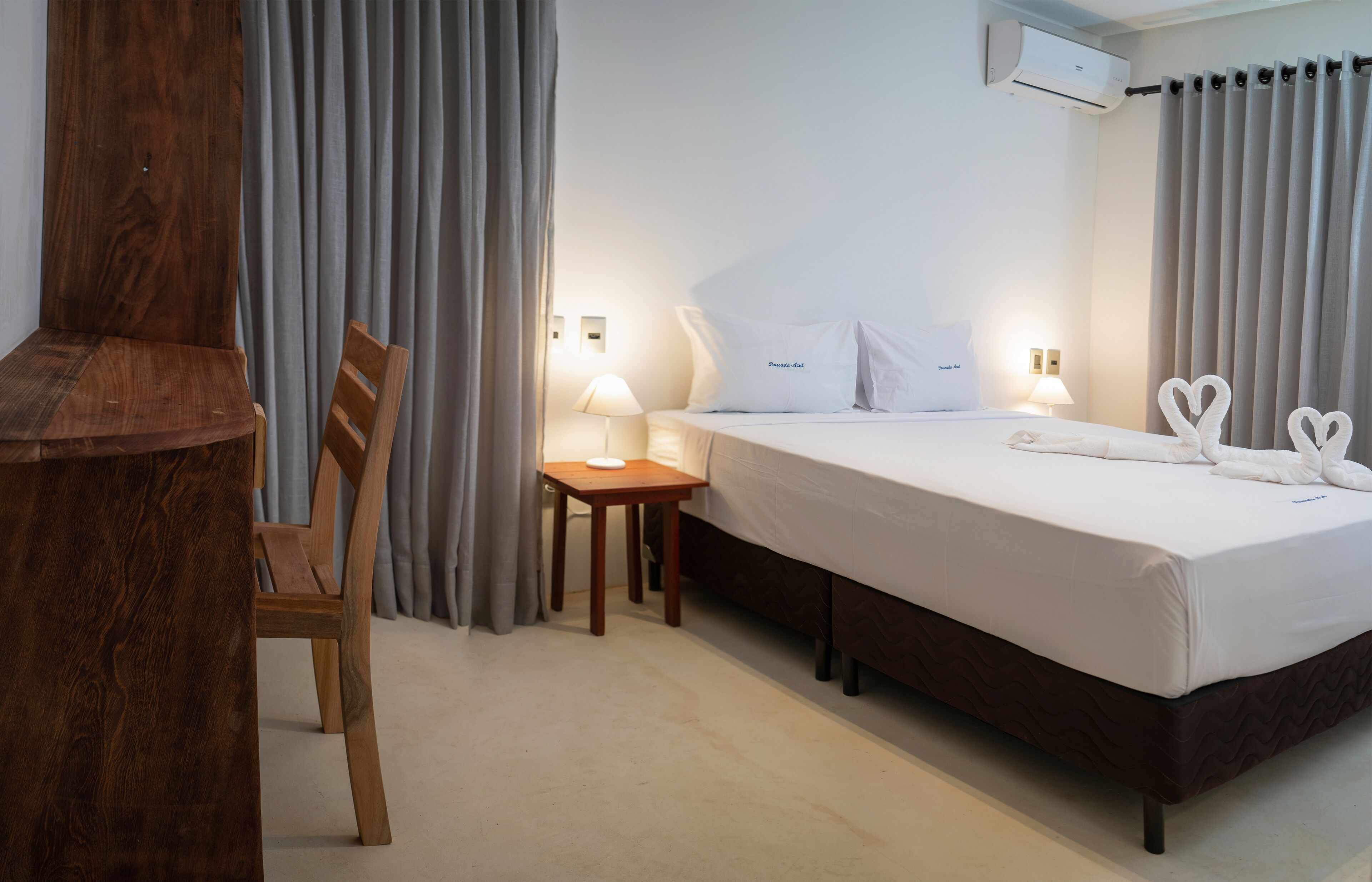 Quarto de hotel com uma cama, uma cadeira de madeira, uma mesinha e uma escrivaninha.