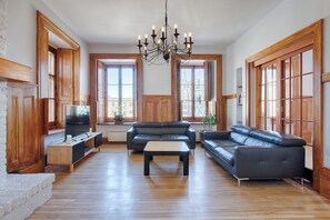 Uma sala de estar ampla com uma TV grande, sofás de couro, piso de madeira e janelas grandes.