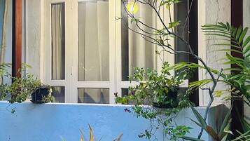 Uma casa com parede externa azul, uma janela com cortina e vários vasos de plantas.