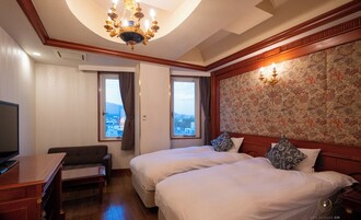 Quarto de hotel com duas camas, uma escrivaninha, uma cadeira, uma televisão e um candelabro.
