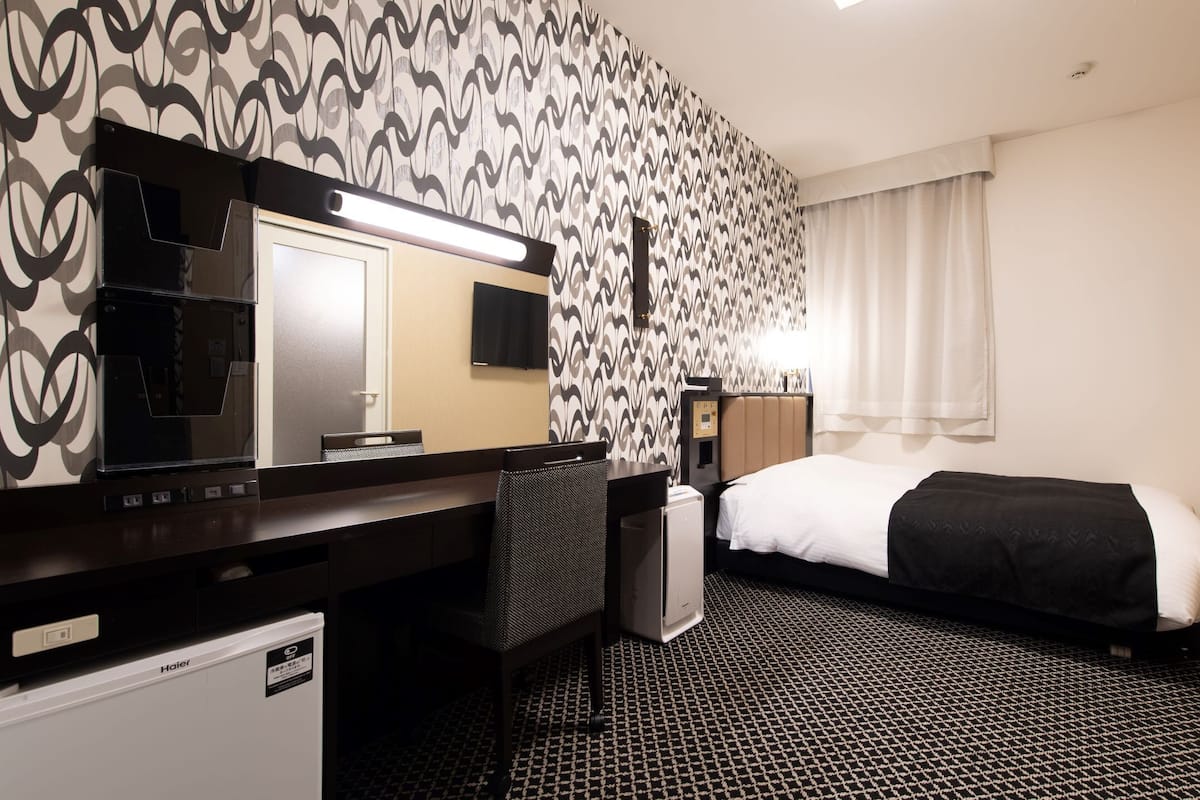 Quarto de hotel com cama, escrivaninha, cadeira, espelho, televisão e painel de controle na parede.