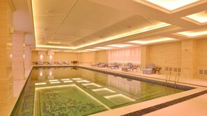 Uma grande piscina coberta com estrutura de vidro, cercada por espreguiçadeiras e teto alto com iluminação embutida.