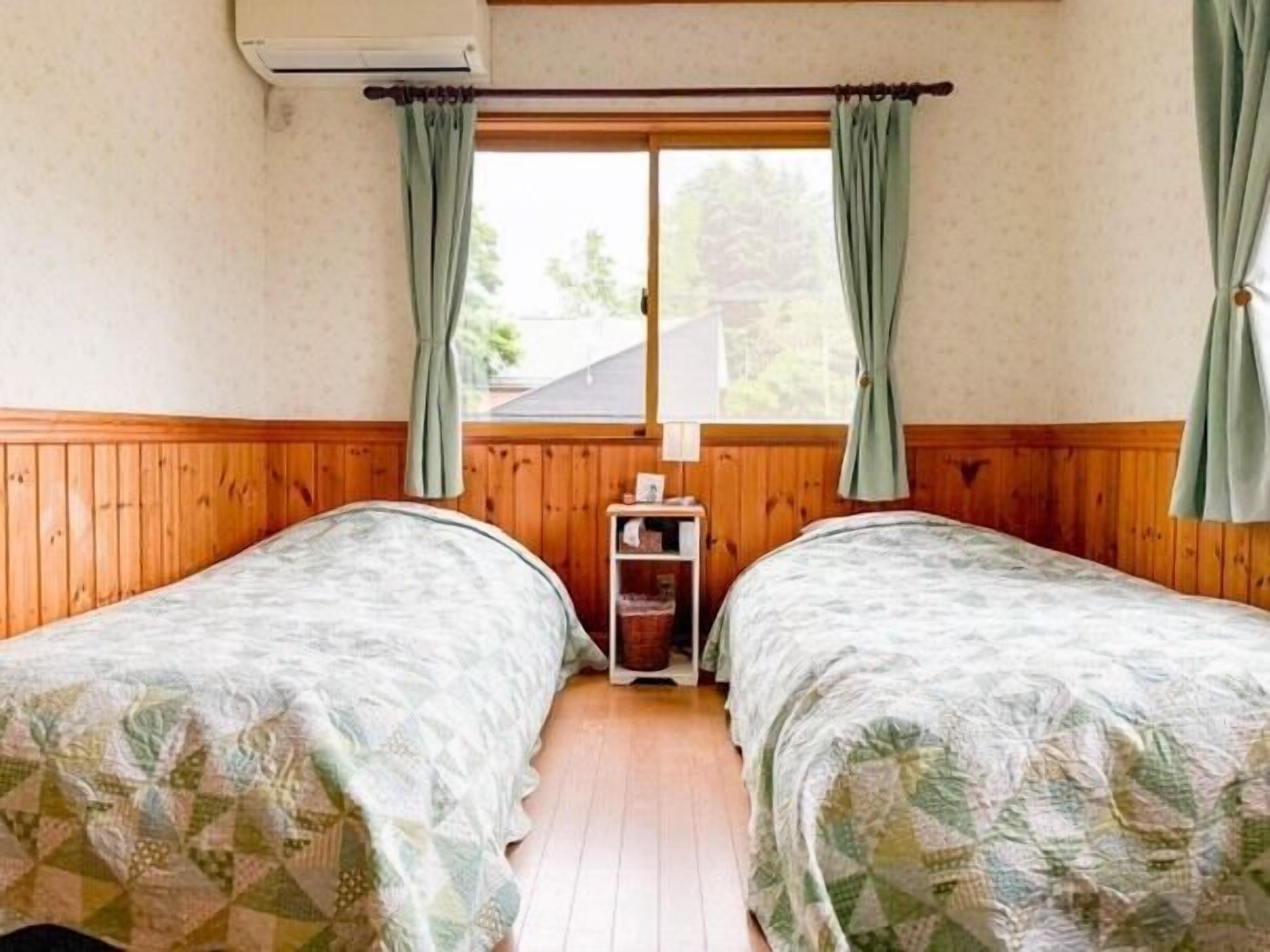 Quarto com duas camas, paredes de madeira, janela com cortinas e uma pequena mesa de cabeceira com abajur.