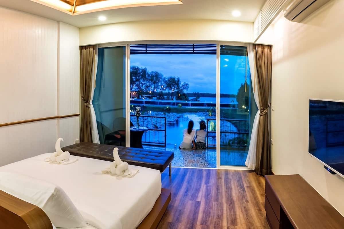Quarto de hotel com cama, televisão, varanda e vista para um espelho d'água e árvores.
