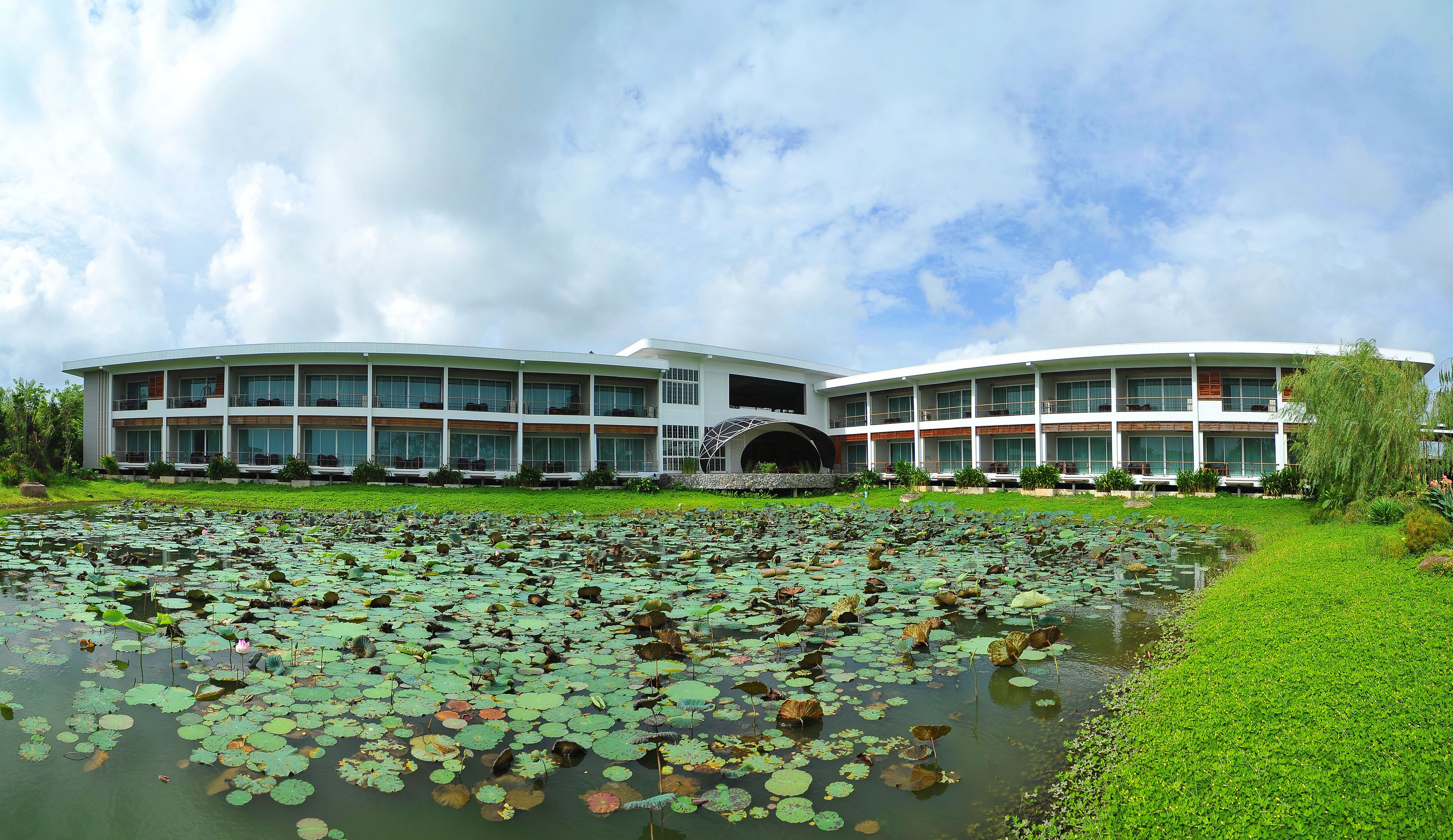 Um edifício moderno com um arco central, rodeado por um lago com folhas de nenúfar e vegetação.