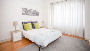 Quarto moderno com cama, mesas de cabeceira, abajur, pintura e cortinas leves.