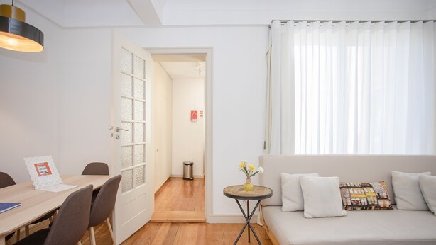 Um quarto com uma mesa de jantar, cadeiras, um sofá, uma mesinha com um vaso e uma janela com cortinas.
