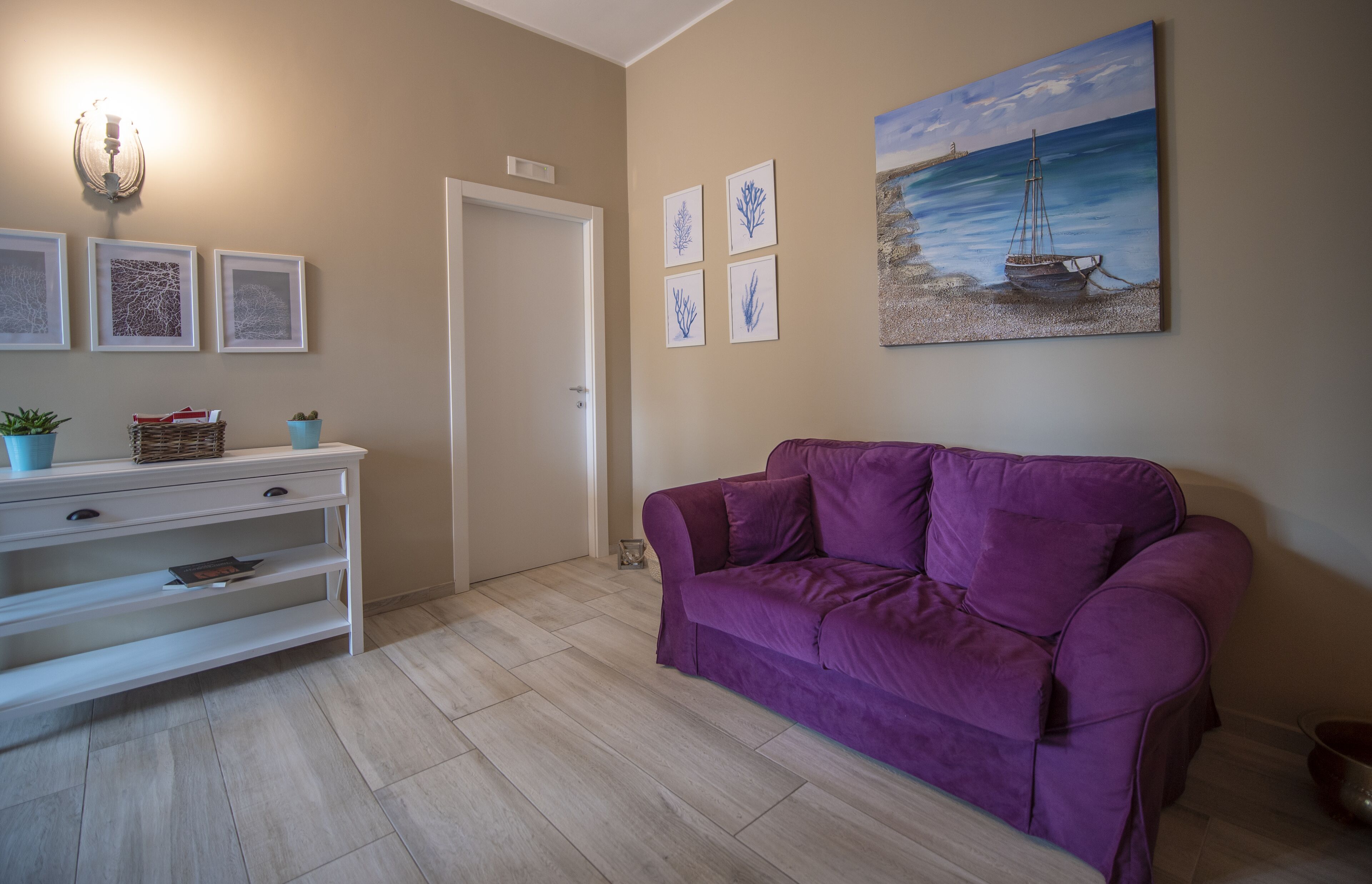 Um quarto com um sofá roxo, uma mesa consola branca com objetos decorativos, e um quadro de um barco na parede.