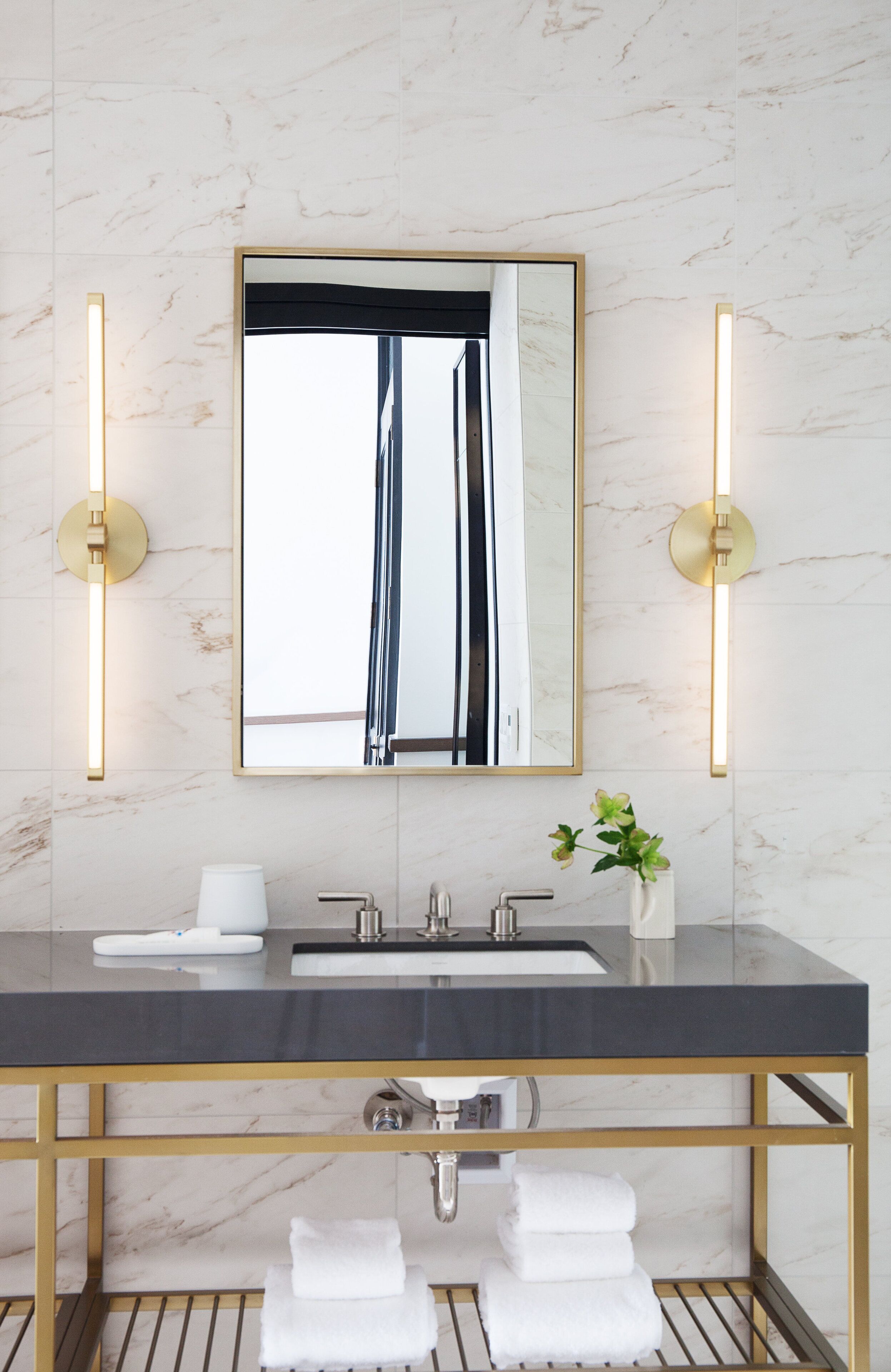 Um banheiro moderno com parede de mármore, pia com bancada preta, detalhes dourados e espelho com iluminação vertical.